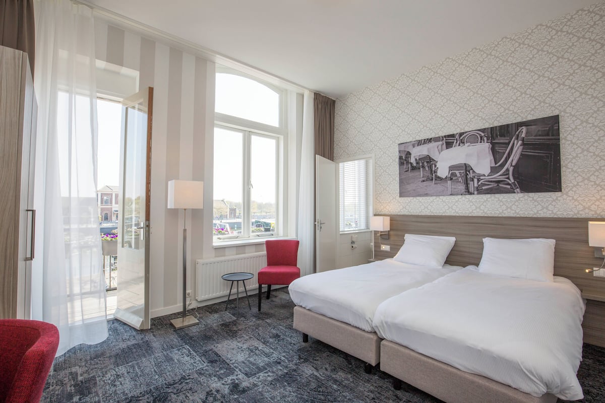 Quarto de hotel com uma cama grande, uma cadeira vermelha, uma mesa pequena e uma janela com cortinas.