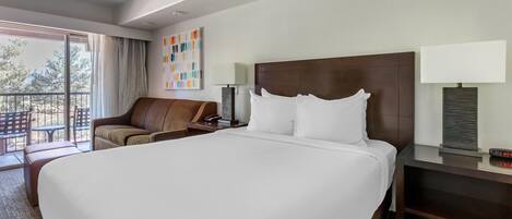 Quarto de hotel com cama grande, escrivaninha com luminária, cadeira e vista para o exterior.