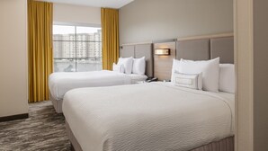 Quarto de hotel com duas camas, uma janela grande com cortinas, cabeceira com luz e piso coberto de carpete.