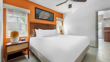Uma cama grande com roupa de cama branca, uma mesa de cabeceira de madeira com abajur, e um ventilador de teto em um quarto com paredes laranja e uma janela grande.