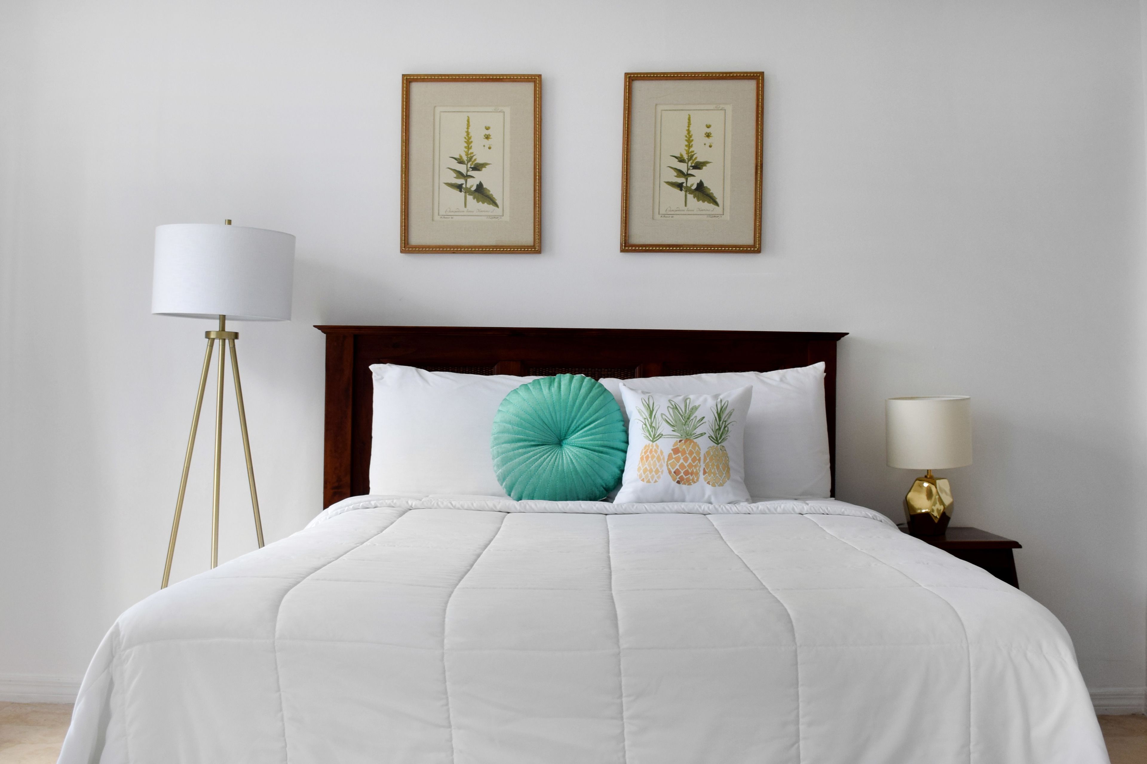 Cama bem arrumada com edredom branco, almofada de detalhes azul-esverdeada e almofadas com tema de abacaxi. Duas gravuras botânicas emolduradas na parede acima da cama.