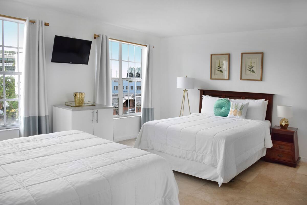 Quarto de hotel com duas camas, televisão presa à parede, escrivaninha com abajur e janela com cortinas.