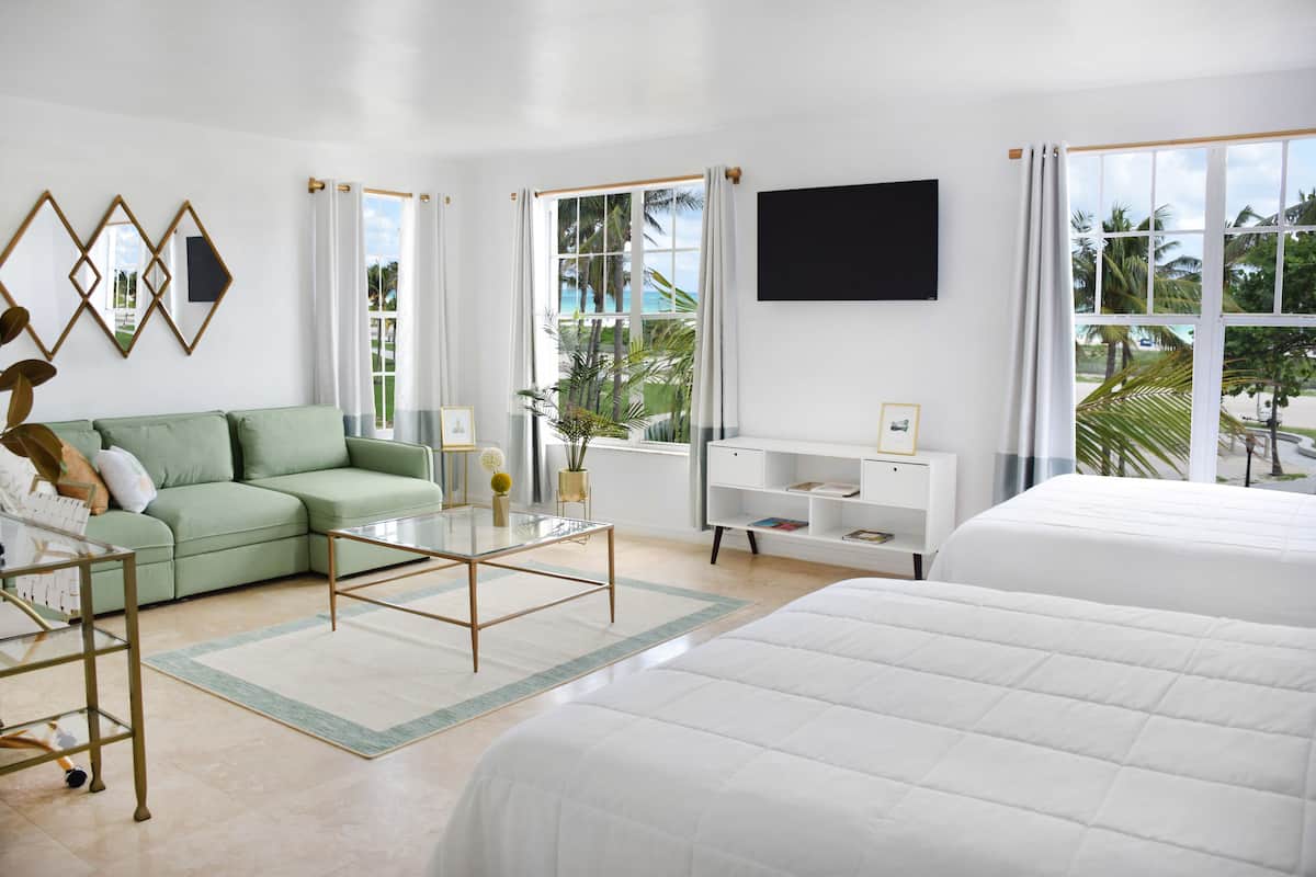 Quarto de hotel moderno com sofá verde, mesa de centro de vidro, cama branca e grandes janelas com vista para palmeiras.