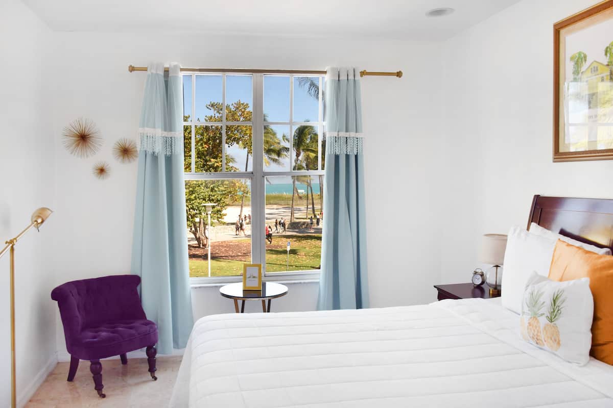 Quarto com cama grande, uma cadeira, mesa de cabeceira, abajur e vista para uma praia e palmeiras através de uma janela.