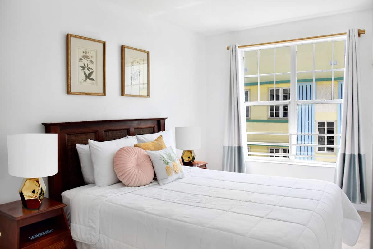 Cama bem arrumada com roupa de cama branca e almofadas decorativas, cabeceira de madeira e mesa de cabeceira com abajur. Há duas gravuras botânicas emolduradas na parede.