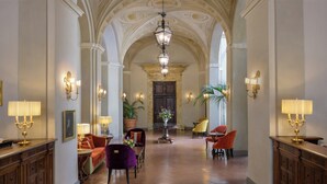 Um hall imponente com pé direito alto, colunas decorativas e um lustre central. Há áreas de estar com mesas e cadeiras, e uma grande planta em vaso no meio.
