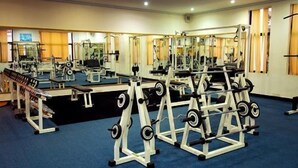 Uma academia com diversos equipamentos de exercício, incluindo máquinas de peso e pesos livres, dispostos em um espaço amplo com carpete azul.