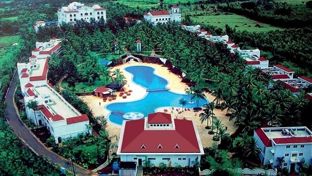 Resort com vários edifícios, piscina central e palmeiras ao redor.
