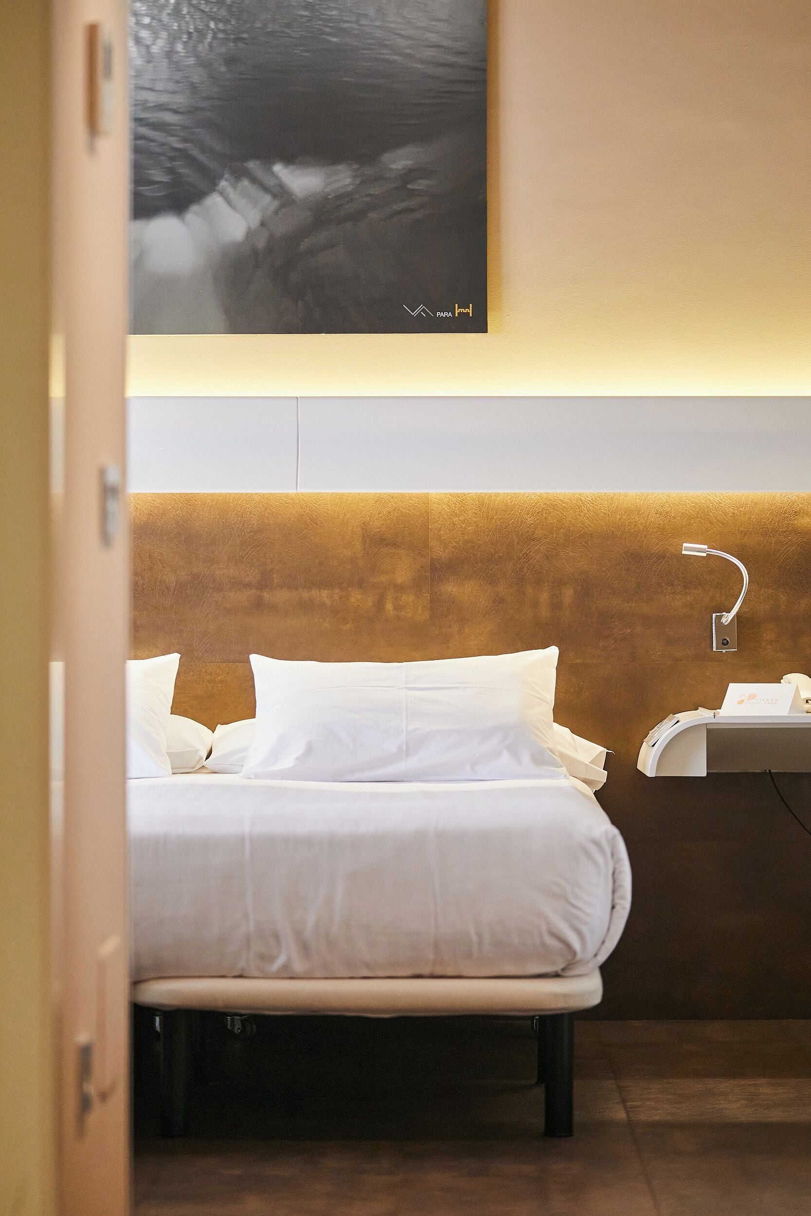 Quarto de hotel moderno com uma cama, mesa de cabeceira, luminária na parede e um quadro emoldurado sobre a cama.