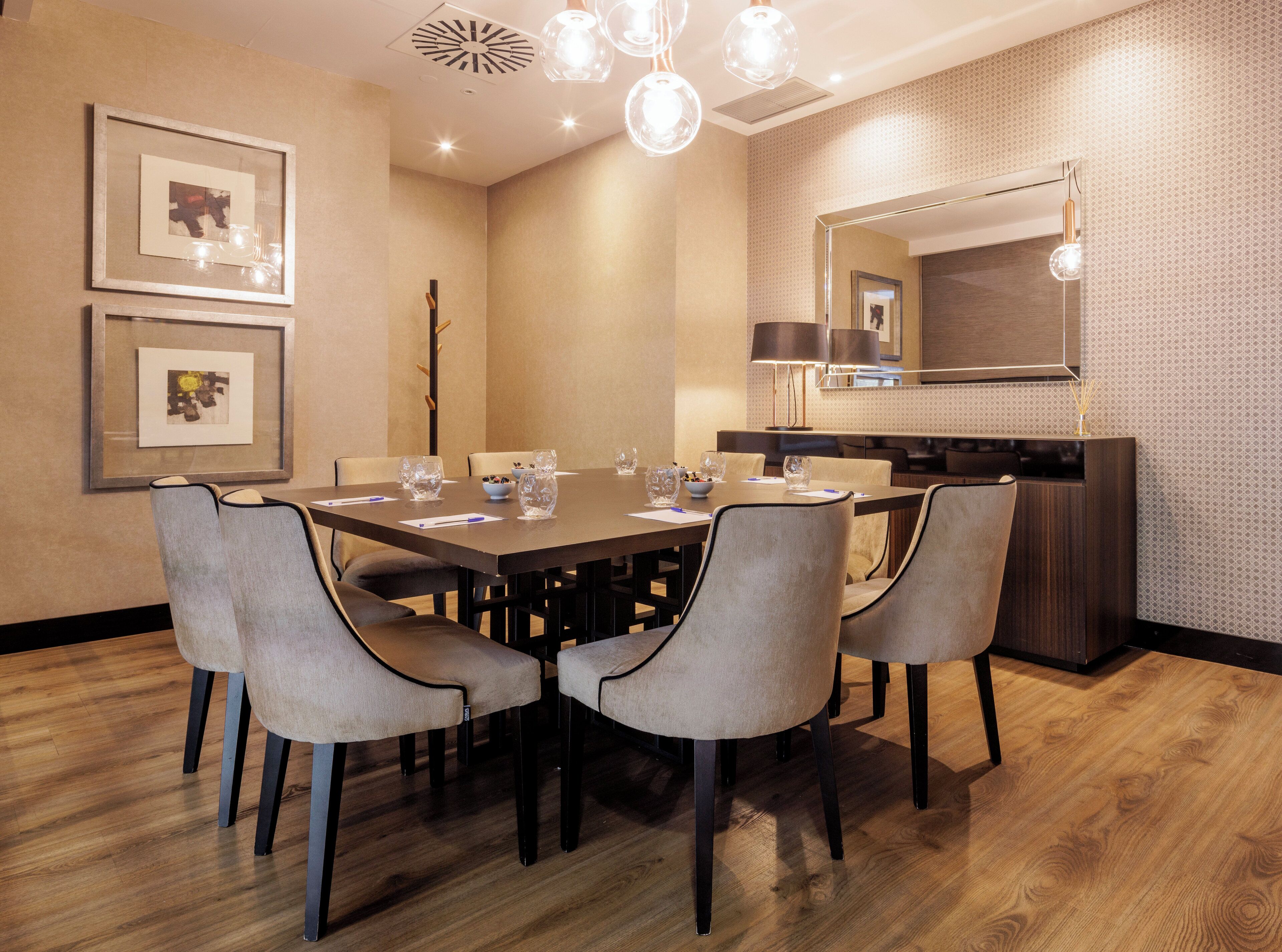 Uma sala de jantar moderna com uma mesa retangular, seis cadeiras, e uma área de cozinha com um espelho grande e luminárias pendentes.