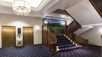 Um saguão de hotel com carpete estampado em azul, um candelabro, uma escadaria com corrimãos de madeira e um mural de uma paisagem.