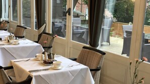 Restaurante com mesas preparadas para refeições, cadeiras e vista da área externa através de grandes janelas.