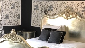 Uma cama com cabeceira elaborada, um banco estofado preto e papel de parede estampado nas paredes.