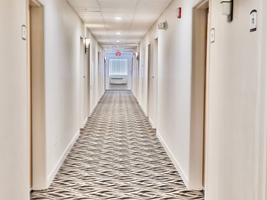 Corredor de hotel com carpete estampado, portas de ambos os lados e um detector de incêndio na parede.