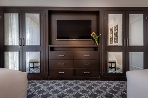 Quarto de hotel com uma grande TV de tela plana fixada em um móvel de madeira escura, uma cama com tapete estampado e um pequeno vaso com flores sobre o móvel.