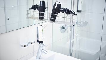 Um banheiro moderno com pia branca, torneira cromada e espelho acima. Dois secadores de cabelo estão fixados numa prateleira de vidro.