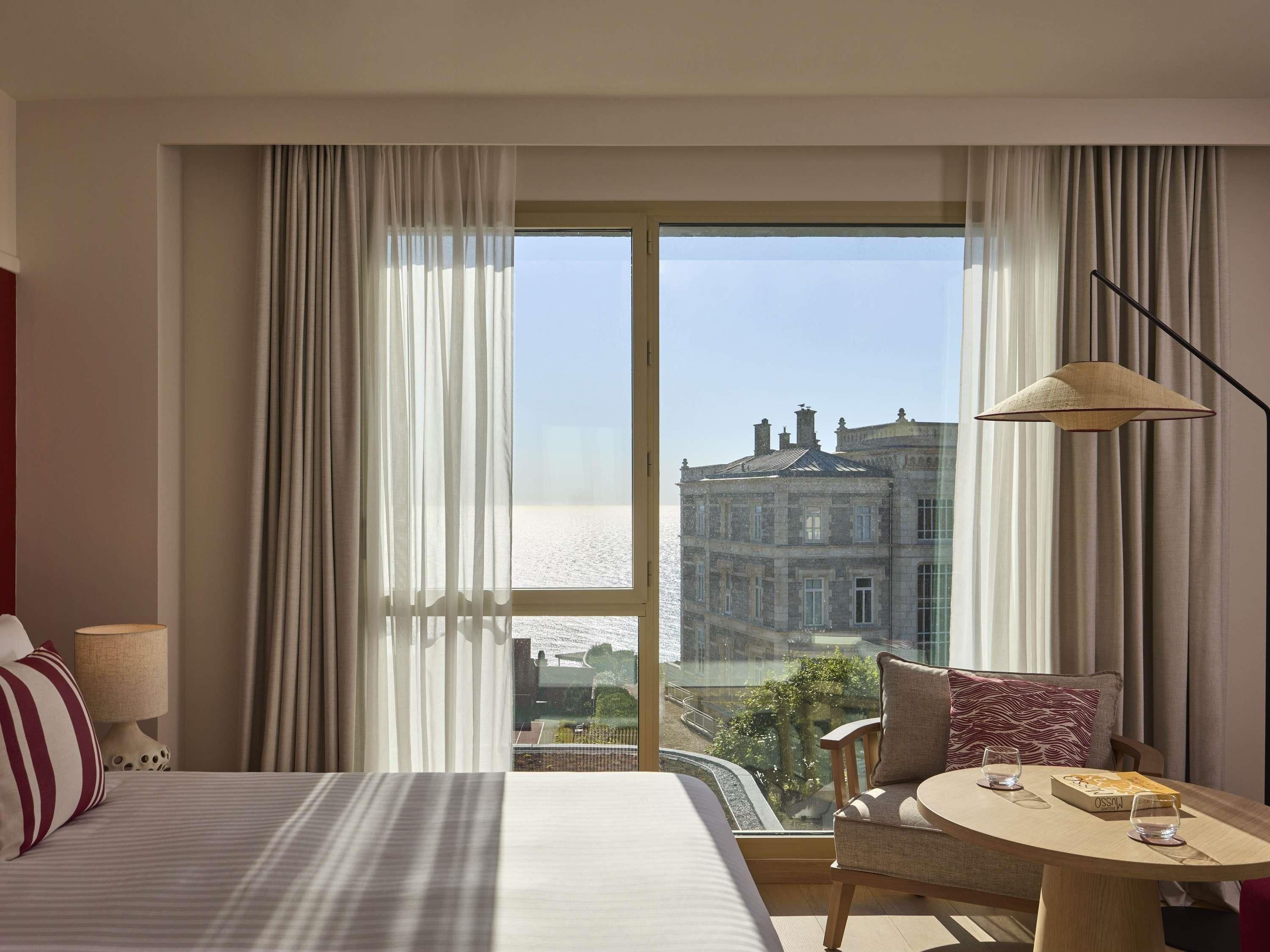 Quarto de hotel com uma janela grande, uma cama, uma mesa redonda com dois copos e um abajur.