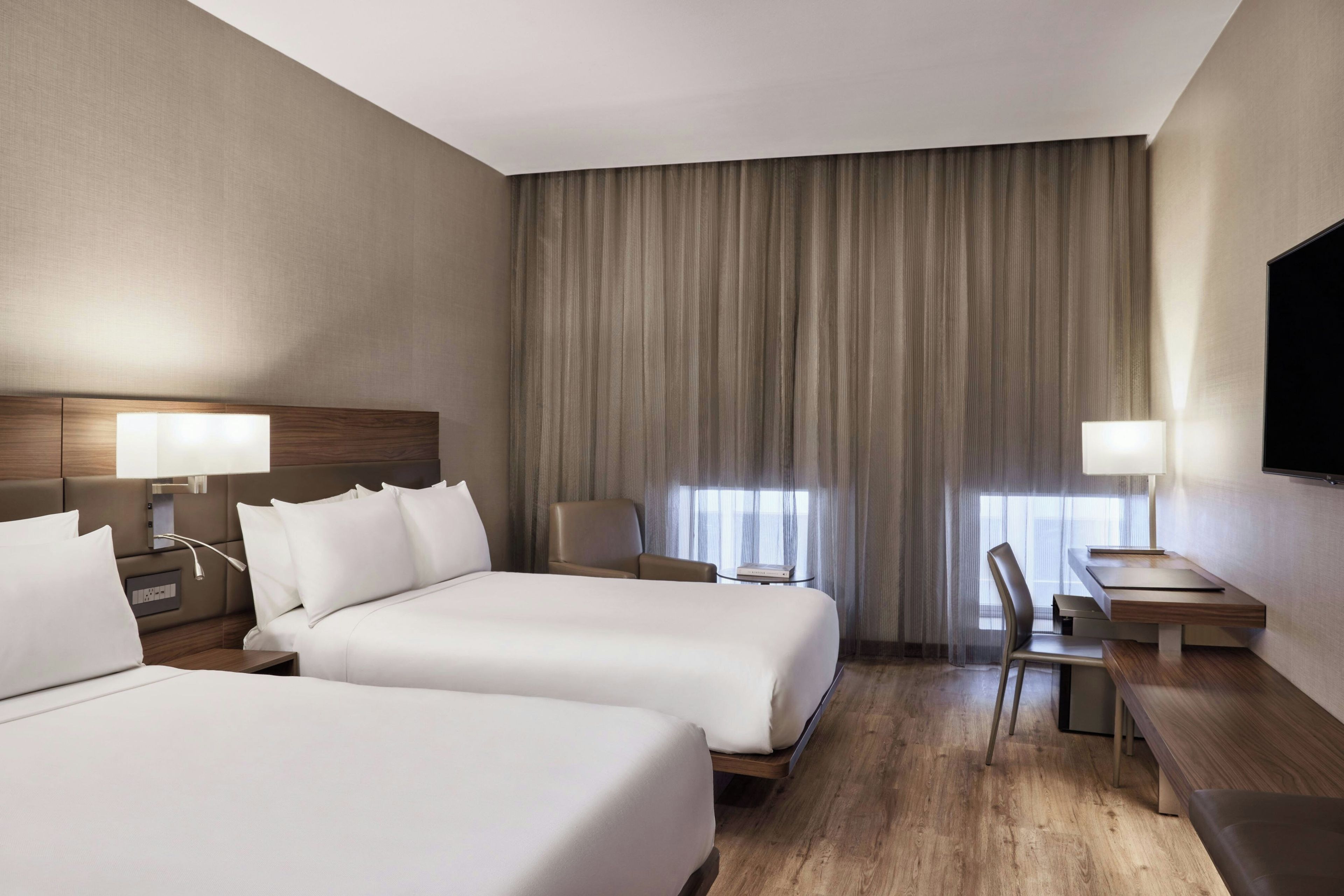 Quarto de hotel com duas camas, uma escrivaninha, uma cadeira e uma televisão.