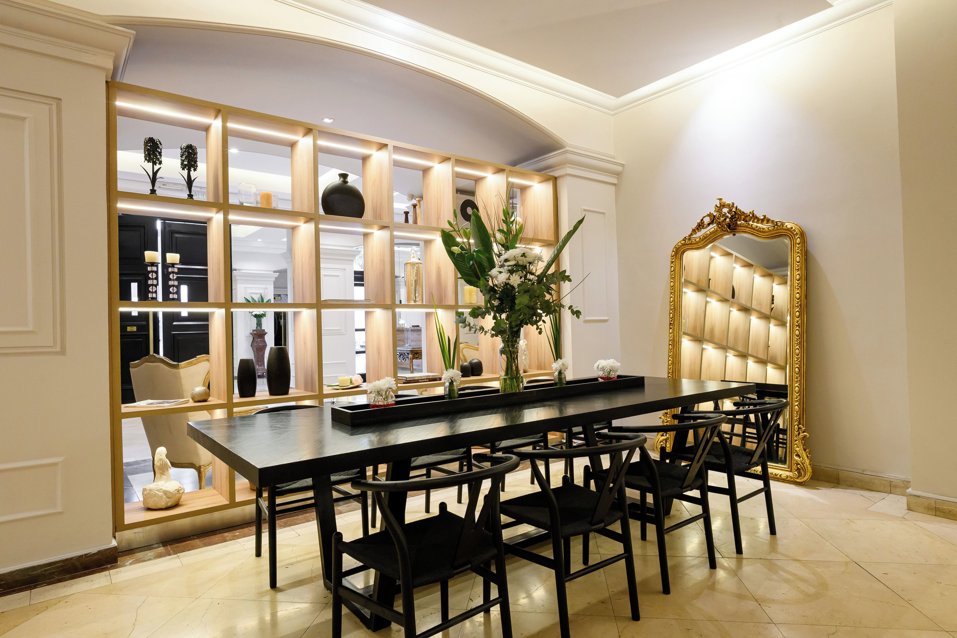 Uma área de refeições com uma mesa comprida e seis cadeiras, um espelho com moldura elaborada e uma estante embutida com diversos objetos decorativos.