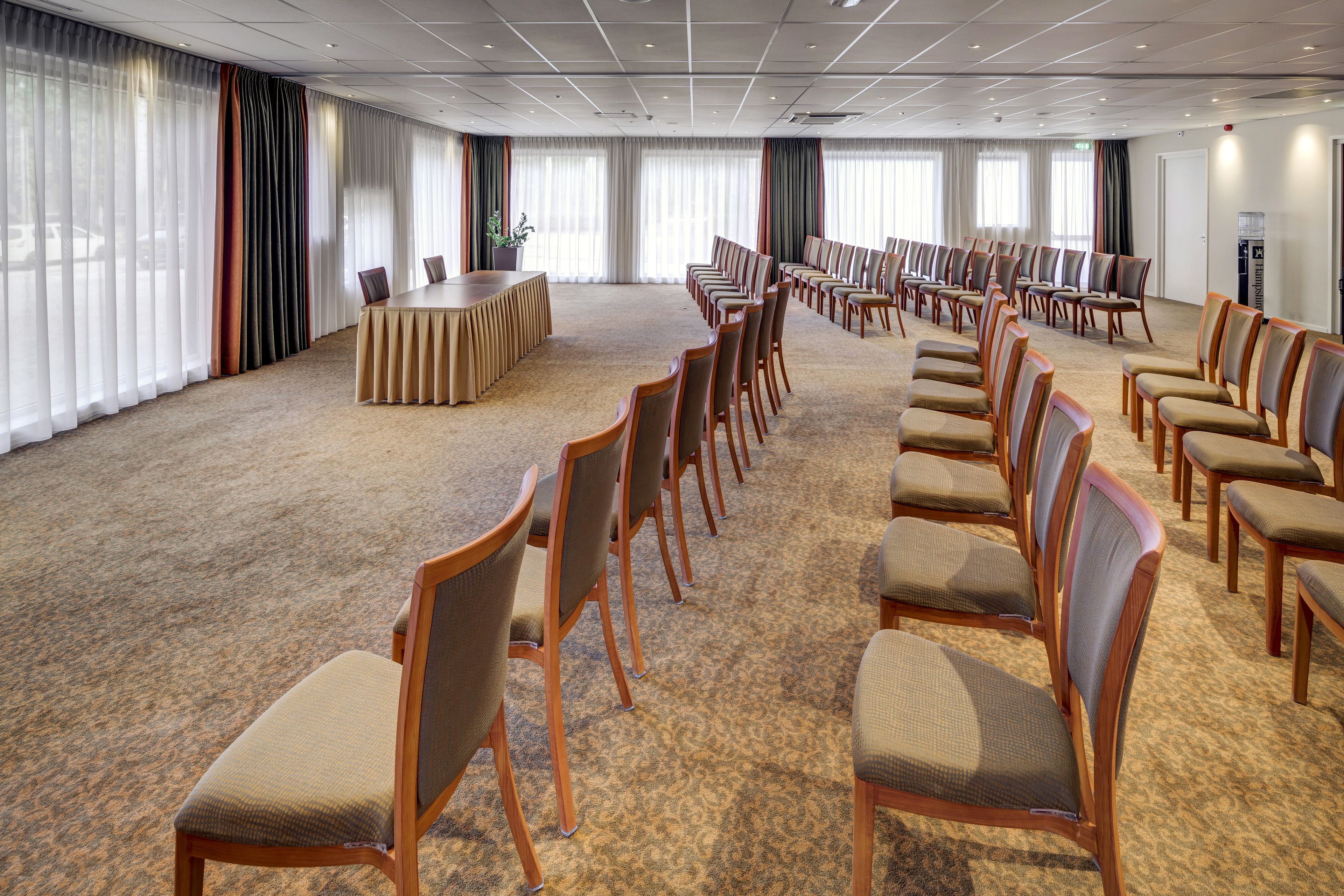 Uma sala de conferências com uma mesa comprida e fileiras de cadeiras, janelas amplas com cortinas e piso coberto de carpete.