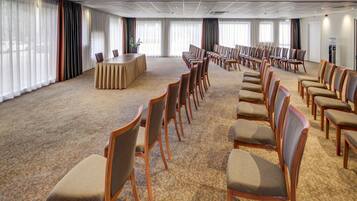 Uma sala de conferências com uma mesa comprida e fileiras de cadeiras, janelas amplas com cortinas e piso coberto de carpete.
