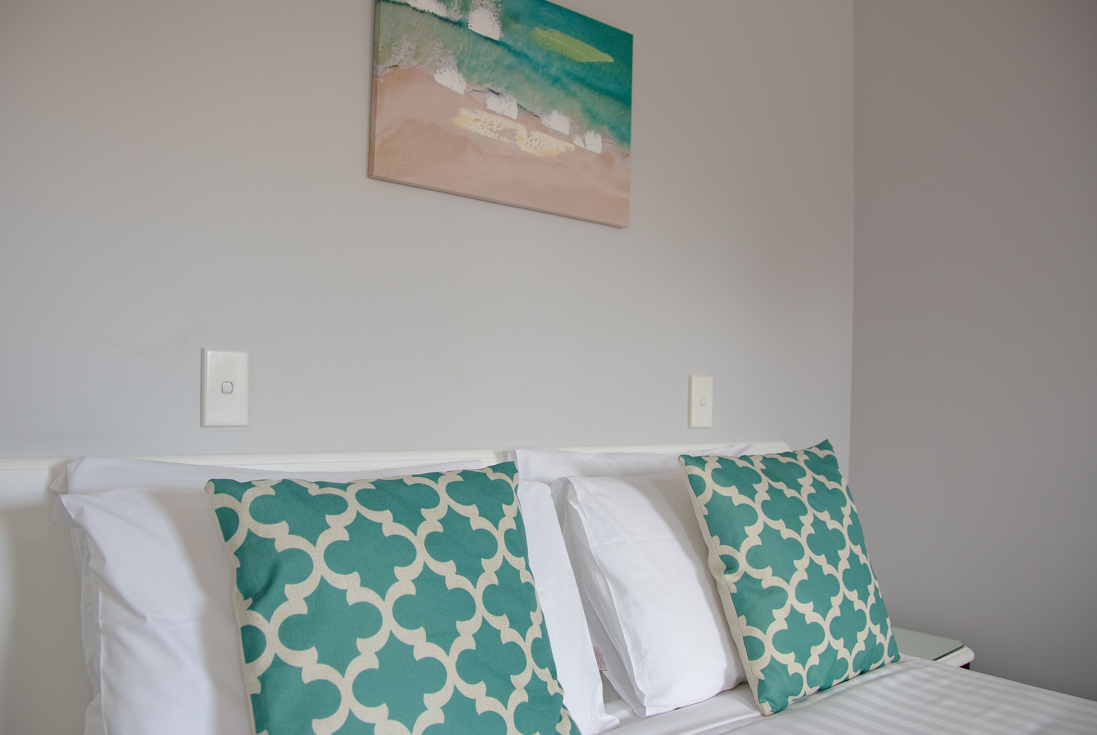 Uma cama com almofadas estampadas em branco e verde-água, uma parede lisa e um pequeno quadro de uma praia.