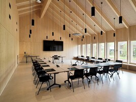 Uma sala de conferência com uma mesa comprida, cadeiras, uma tela grande e luz natural entrando por janelas amplas.