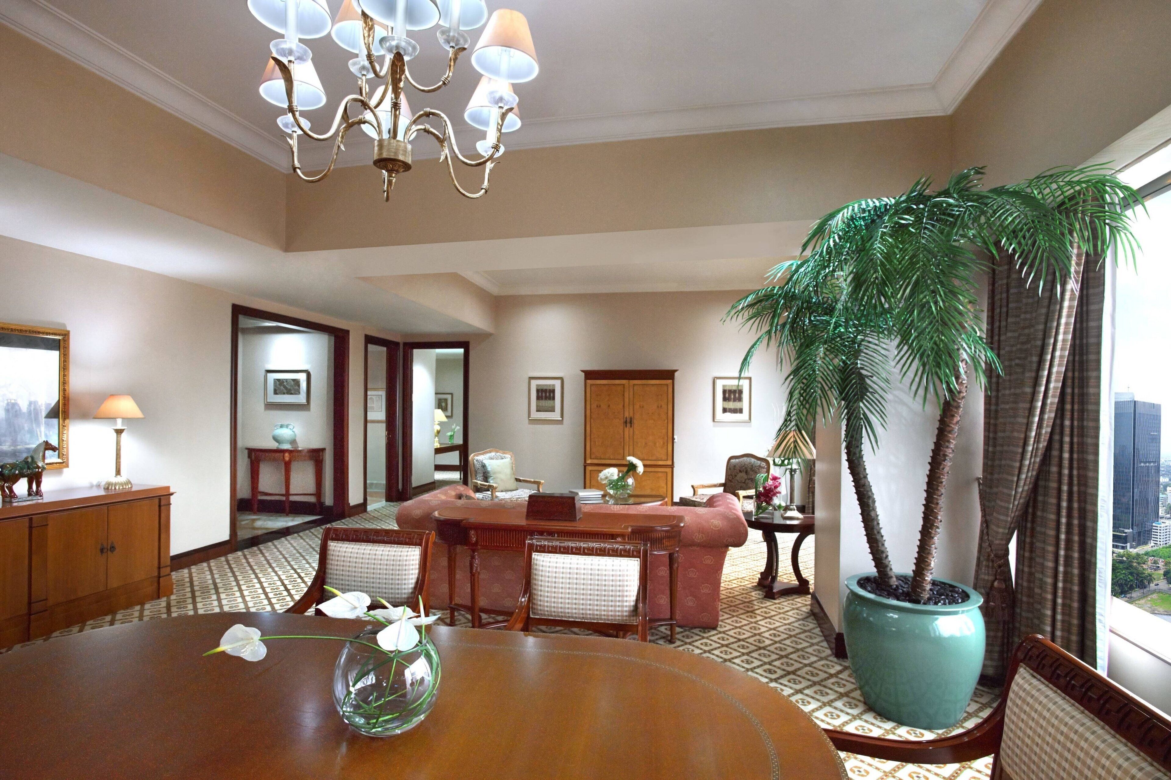 Uma sala de jantar com uma mesa de madeira, cadeiras, um candelabro, uma planta grande em vaso e quadros nas paredes.