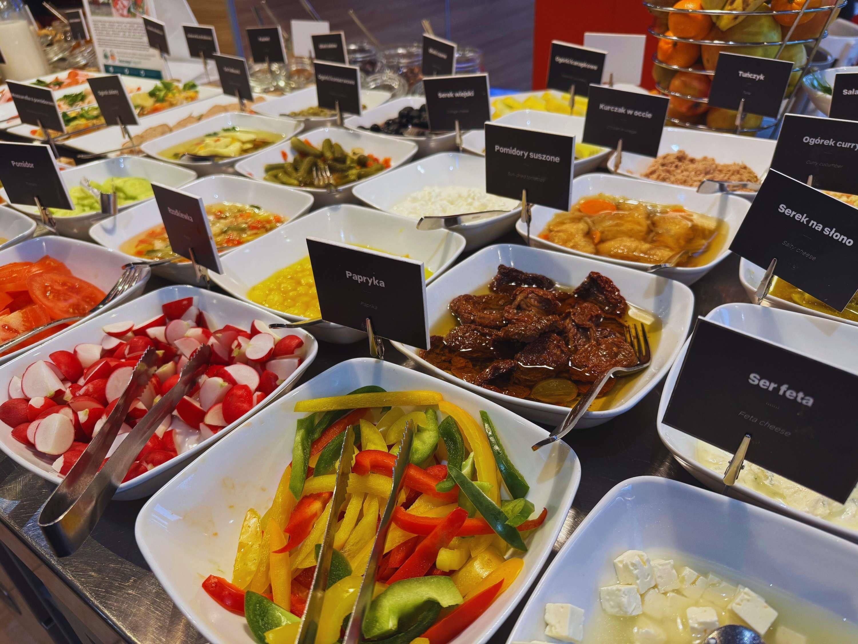 Buffet com diversos pratos identificados em língua estrangeira, incluindo saladas, carnes e vegetais.
