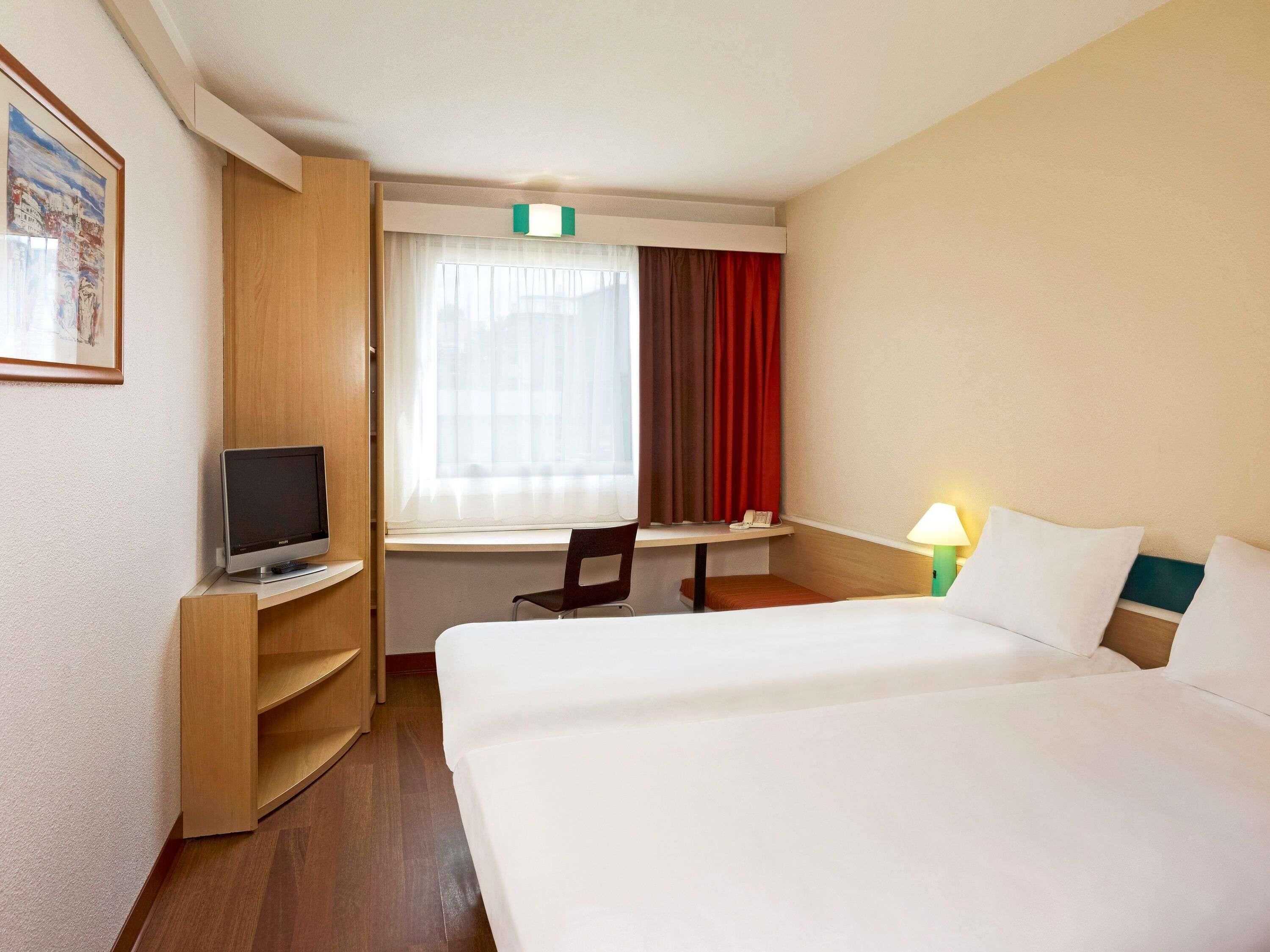 Quarto de hotel com cama, escrivaninha com cadeira, televisão, janela com cortinas e luminária.