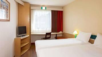 Quarto de hotel com cama, escrivaninha com cadeira, televisão, janela com cortinas e luminária.