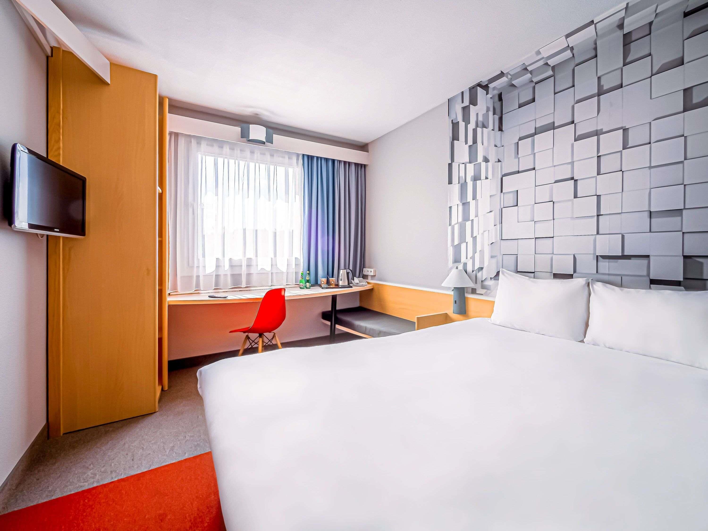 Quarto de hotel com uma cama, uma escrivaninha com cadeira, uma televisão na parede e uma janela com cortinas.