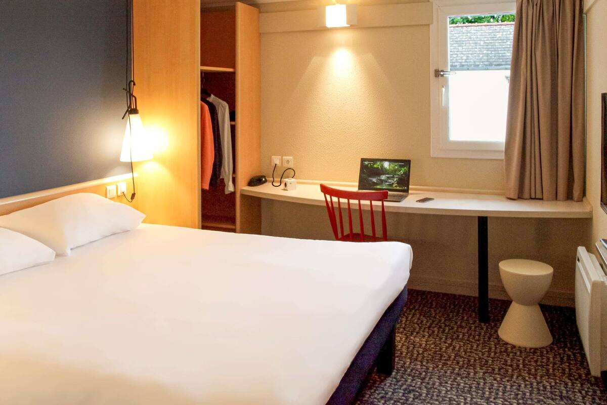 Quarto de hotel com cama, escrivaninha com laptop, cadeira, sanita e guarda-roupa.