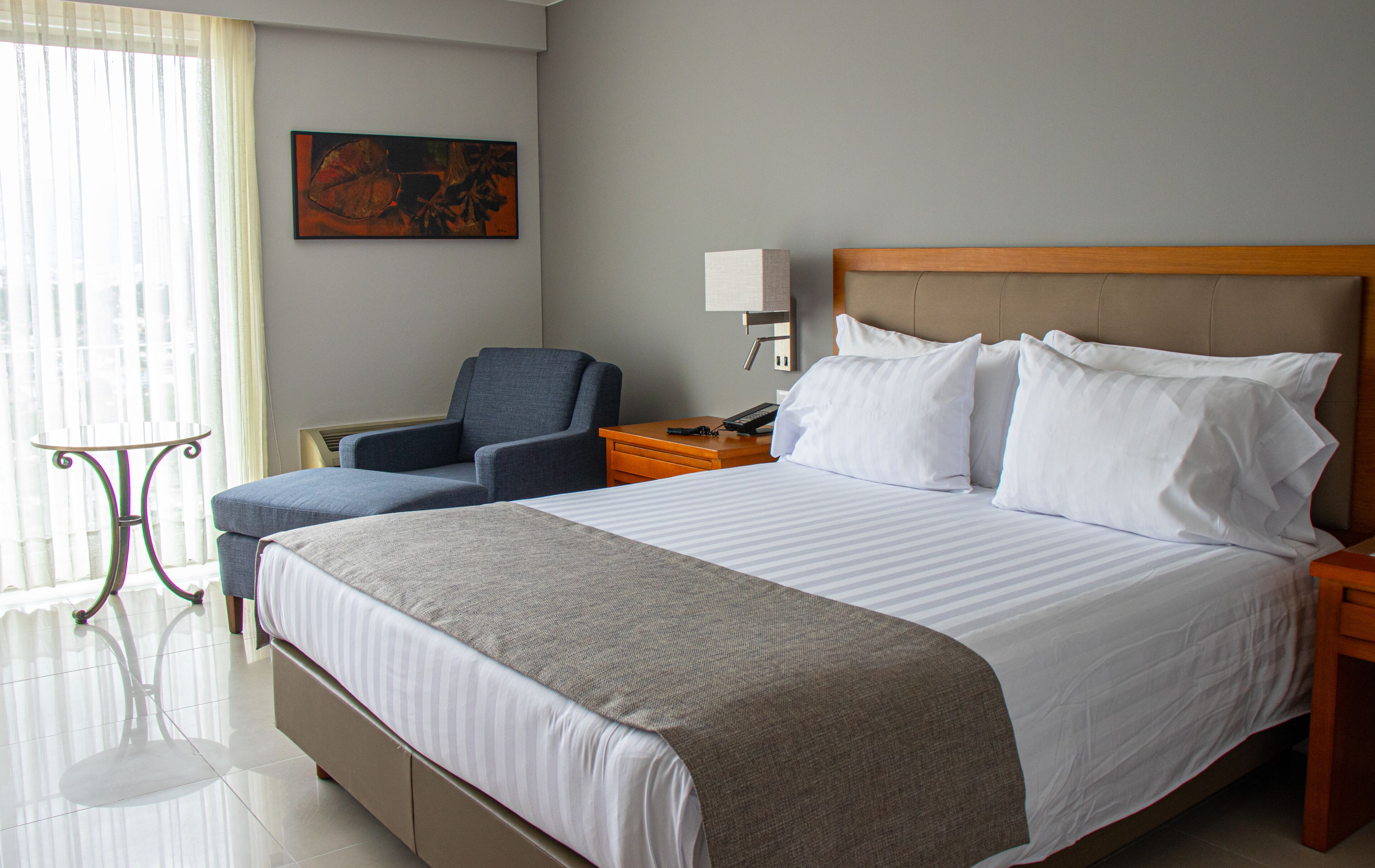 Quarto de hotel com uma cama grande, uma cadeira, uma mesa de cabeceira, um abajur e um quadro na parede.