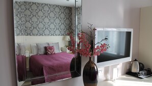 Quarto com um espelho grande, uma cama com colcha rosa, um vaso com flores rosa, uma televisão de tela plana e uma cafeteira no criado-mudo.