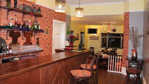 Uma área de bar com parede de tijolos, prateleiras com garrafas e copos, e luminárias pendentes. Há também um espaço de refeições com cadeiras e uma mesa.