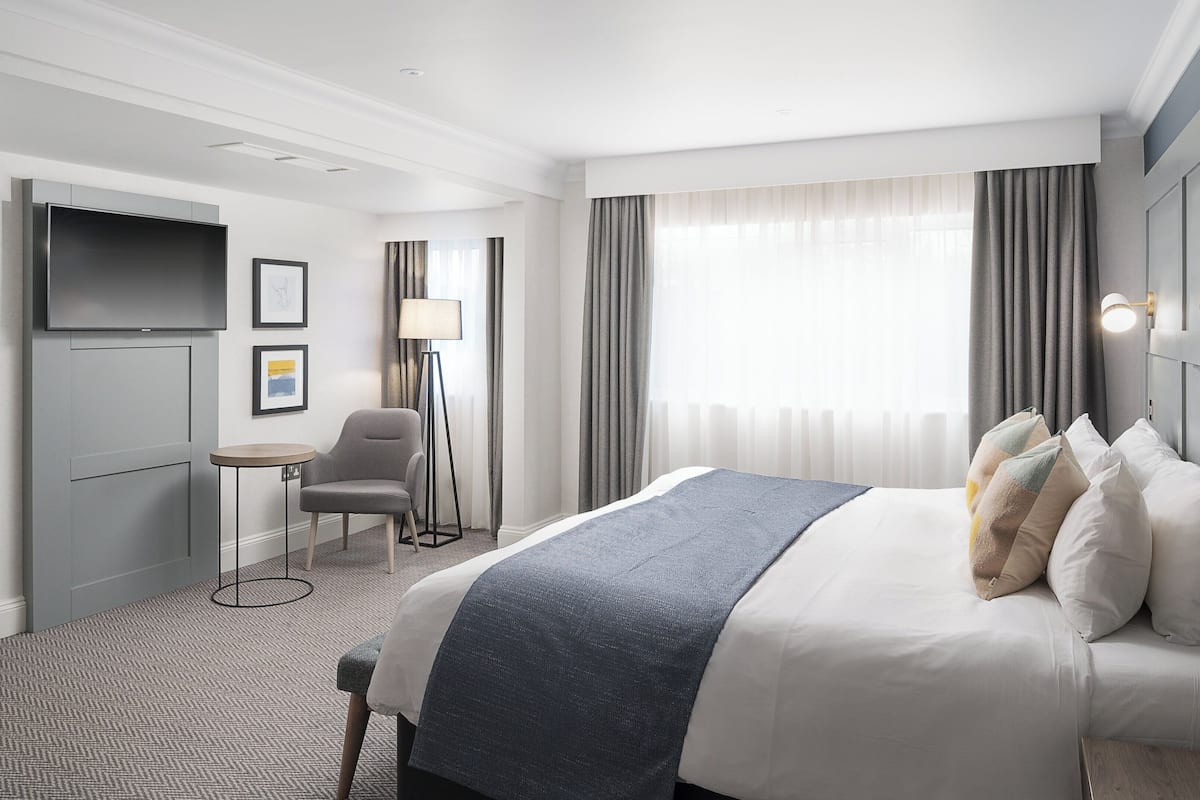 Quarto de hotel com cama, escrivaninha, cadeira, televisão e janela com cortinas.