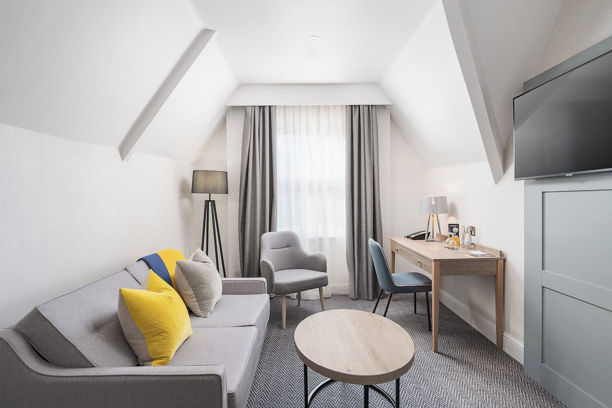 Quarto de hotel moderno com sofá, duas cadeiras, uma escrivaninha de madeira, uma mesa redonda, TV e abajur.