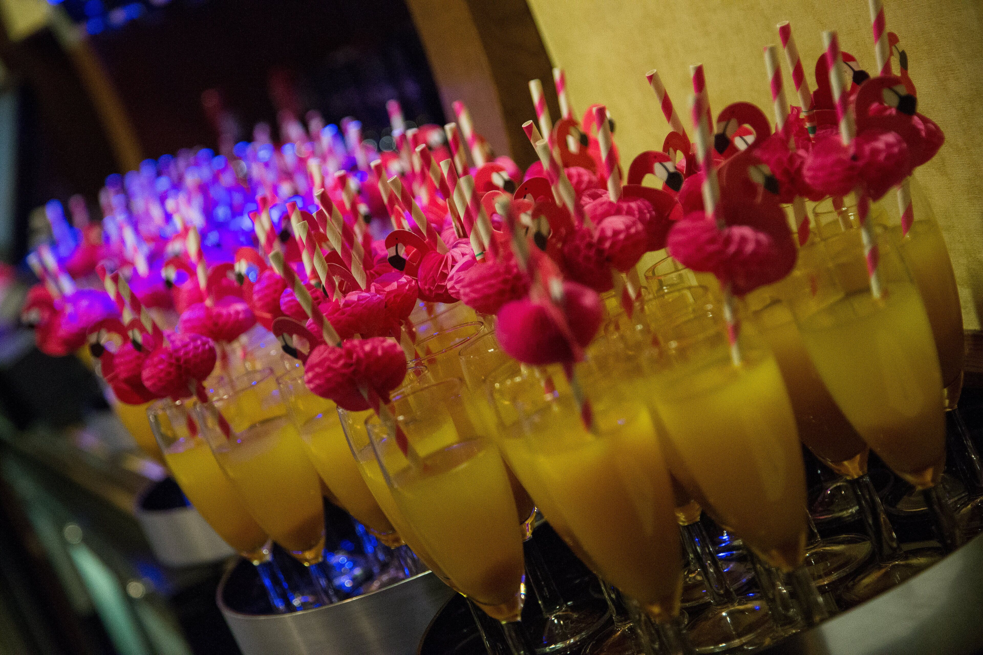 Vários copos com decorações de flamingos cor-de-rosa e canudos, cheios de uma bebida amarela.