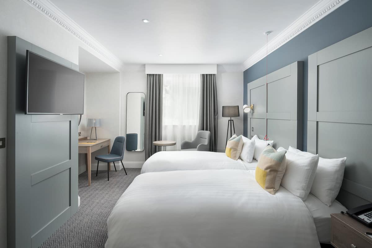 Quarto de hotel com duas camas, uma escrivaninha, uma cadeira, uma televisão e um abajur.