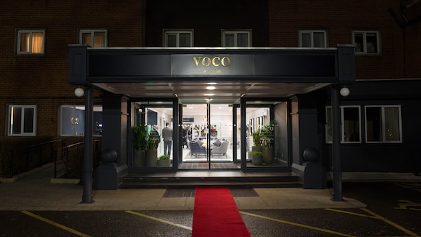 Entrada de hotel com tapete vermelho, portas de vidro e placa com a inscrição "VOCO".