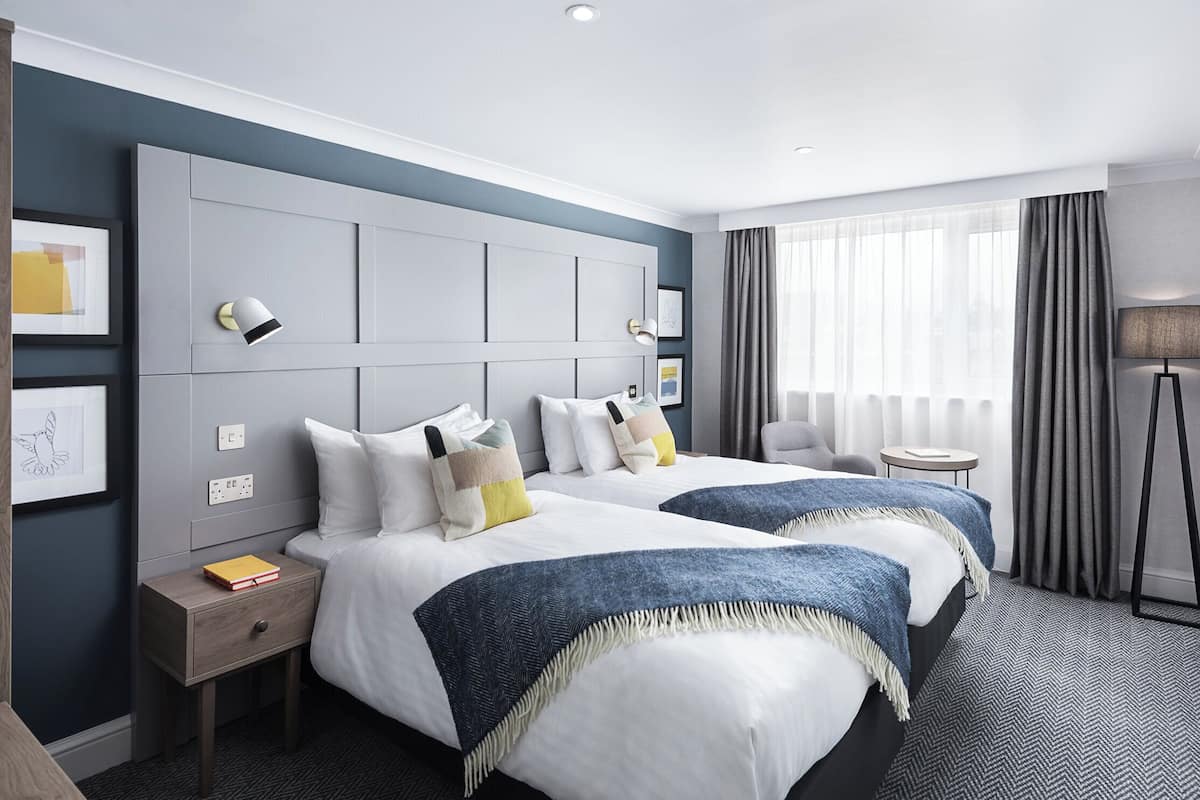 Quarto de hotel com duas camas, uma cabeceira grande, uma mesa de cabeceira com abajur, uma mesinha redonda e um candeeiro de chão.