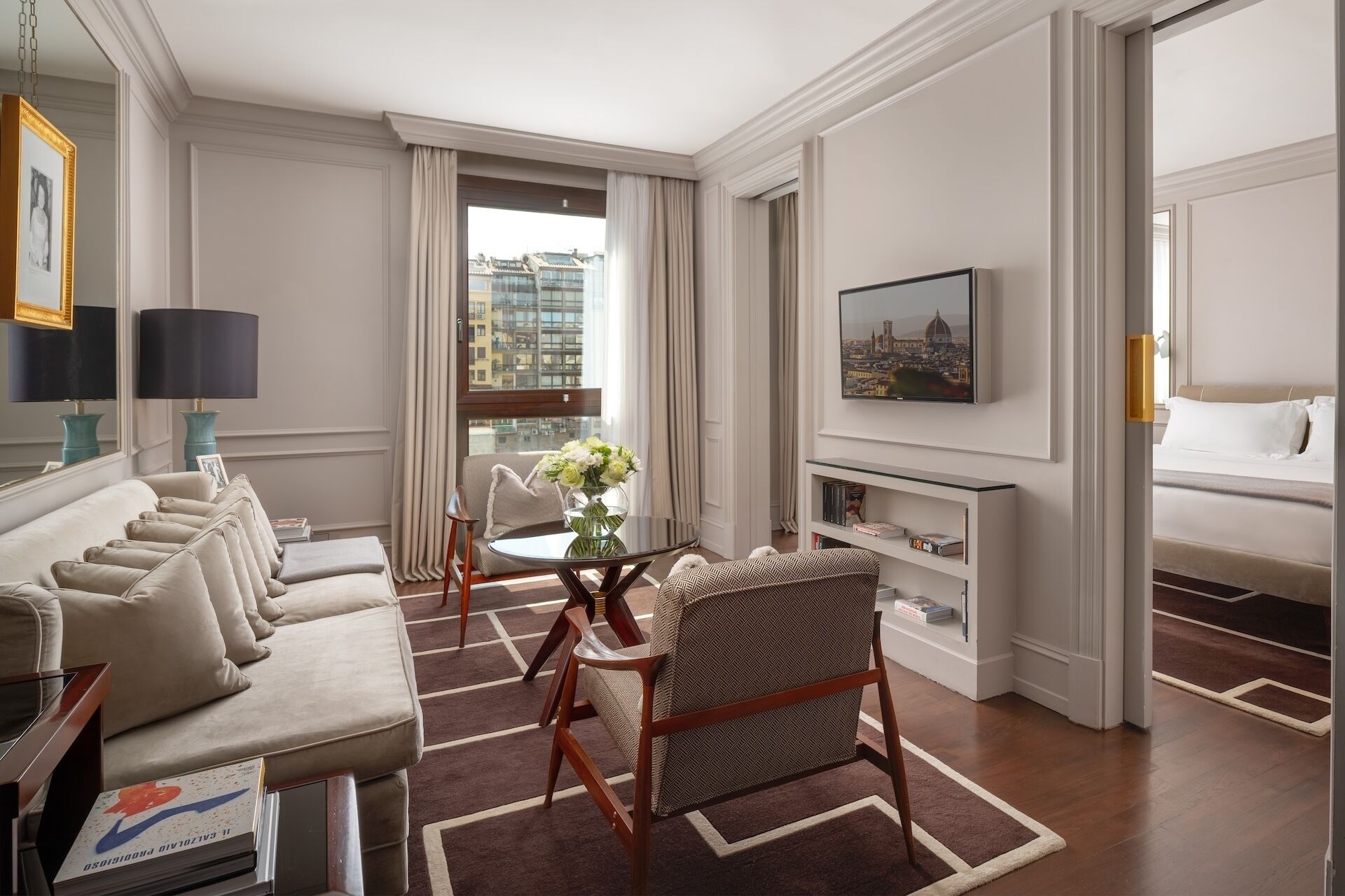 Quarto de hotel moderno com sofá, poltrona, mesa pequena e televisão em suporte.