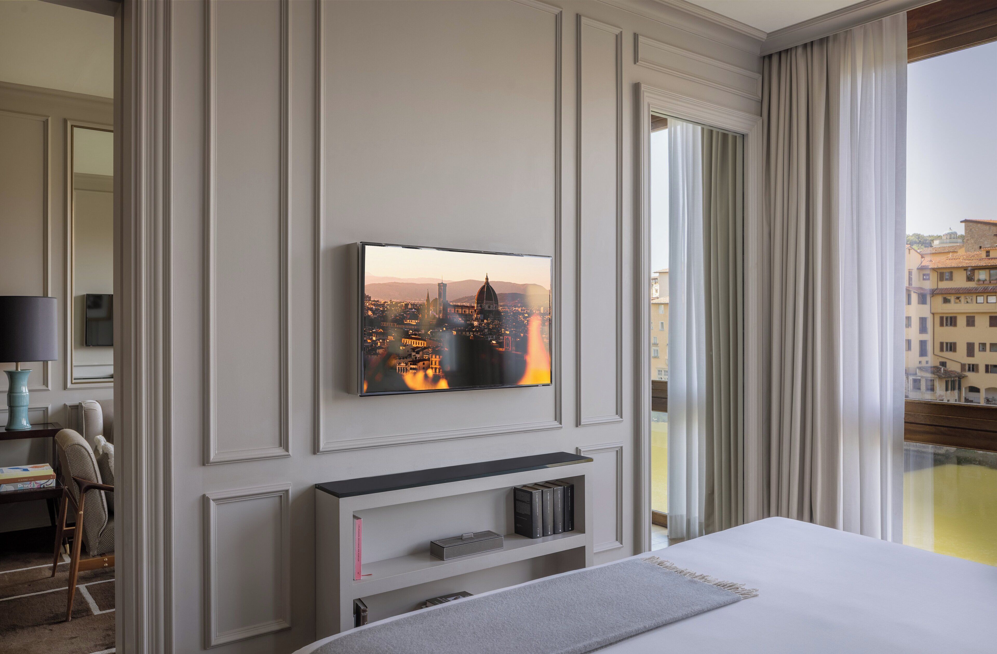 Quarto de hotel com televisão de tela plana na parede, cama com cabeceira cinza, mesa de cabeceira pequena com abajur e vista para a cidade através de amplas janelas.