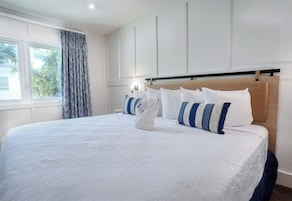 Cama bem arrumada com roupa de cama branca e almofadas listradas em azul, cabeceira de madeira e janela com cortina azul.