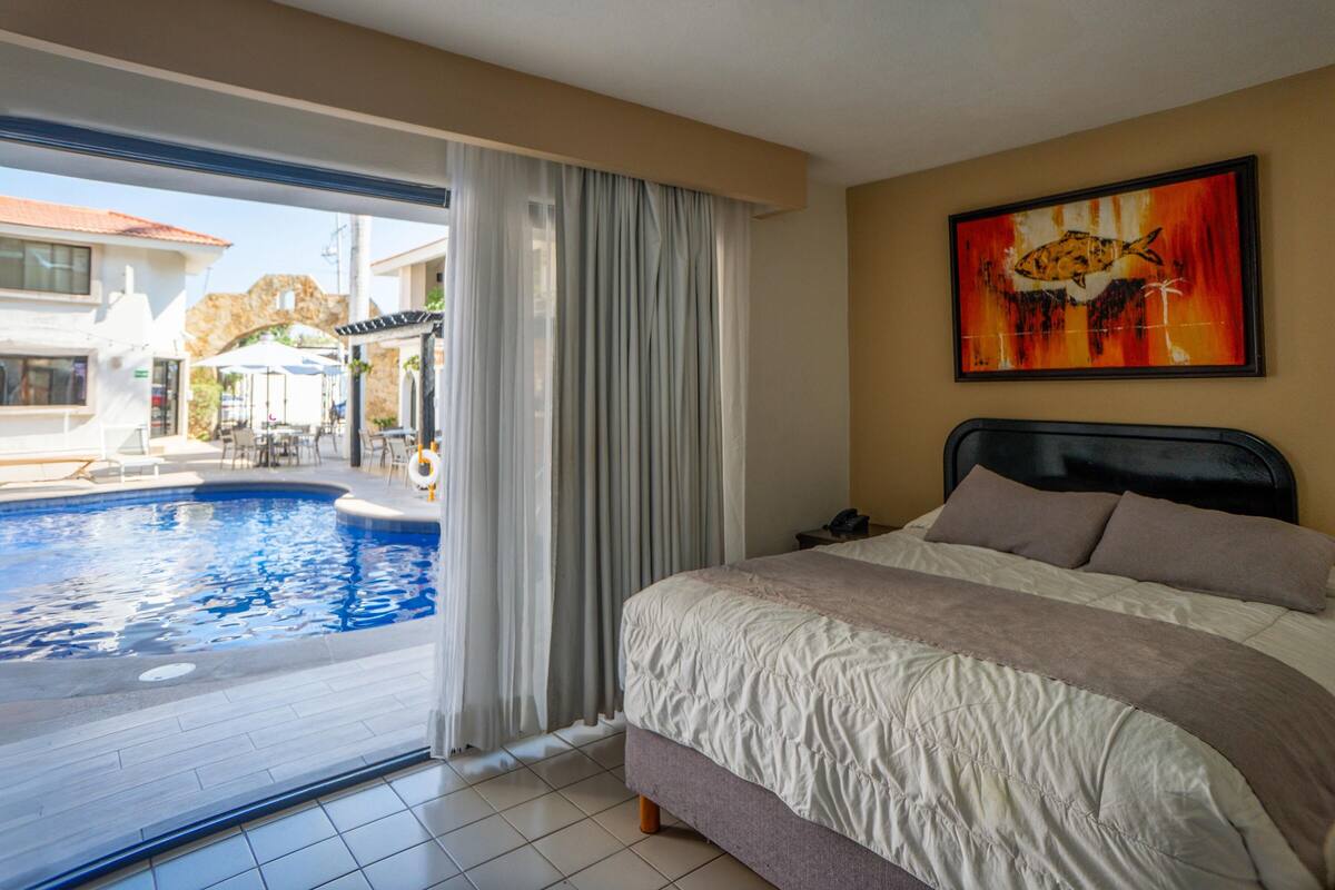 Quarto de hotel com cama, um quadro, vista para piscina ao ar livre e área de varanda com guarda-sóis.