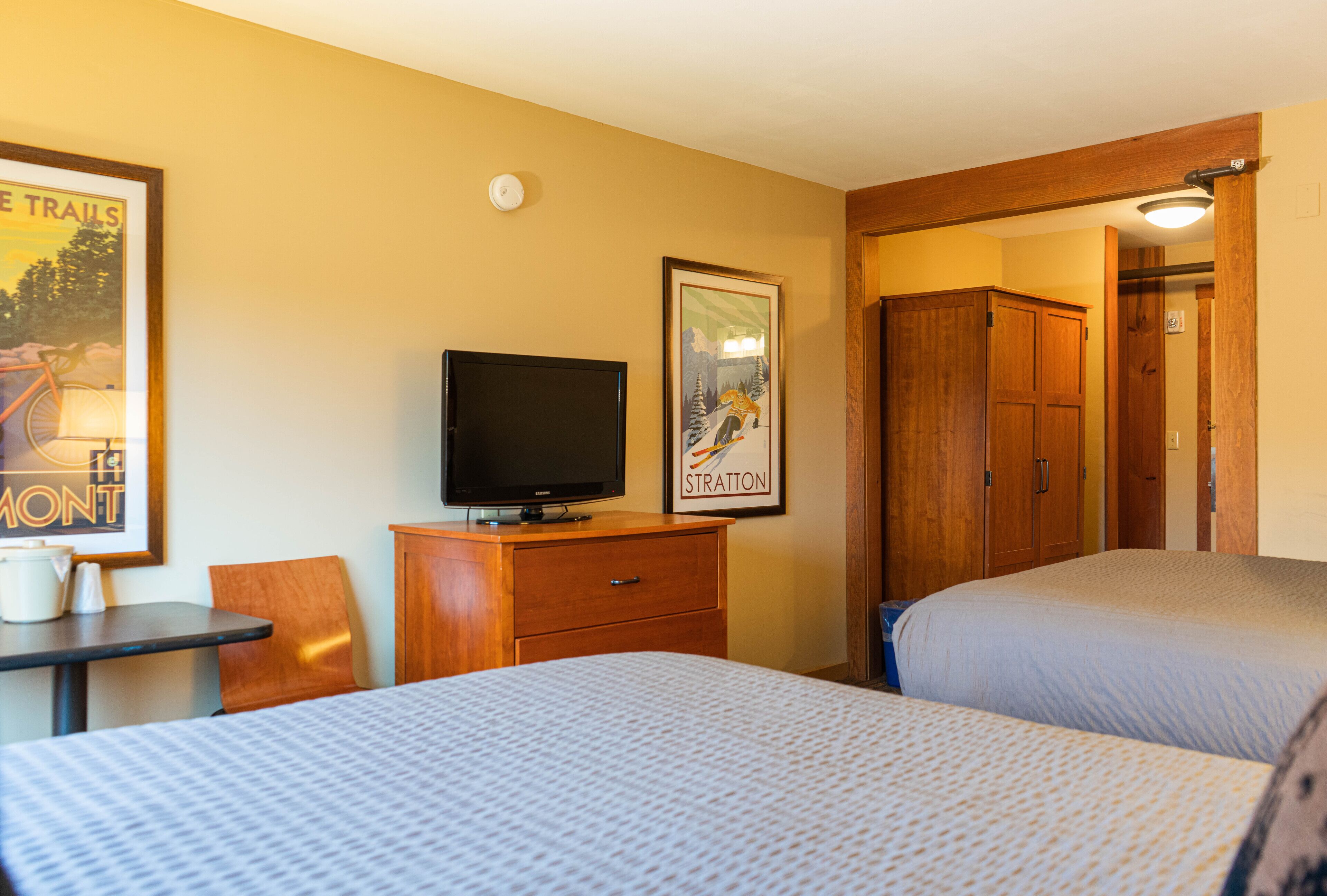 Quarto de hotel com duas camas, uma televisão em uma cômoda de madeira, um quadro na parede e uma porta que dá para outro quarto.