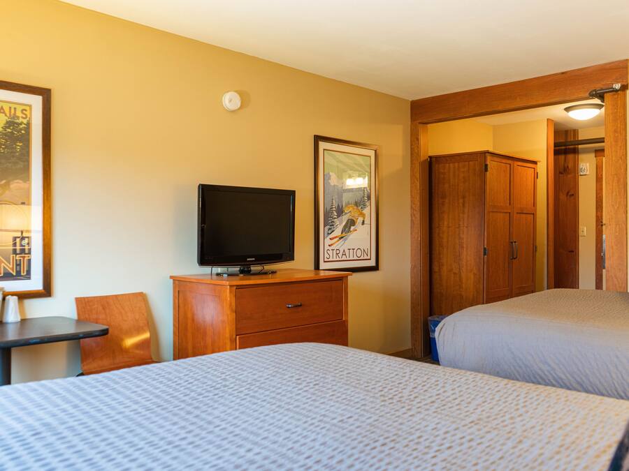 Quarto de hotel com duas camas, uma televisão em uma cômoda de madeira, um quadro na parede e uma porta que dá para outro quarto.