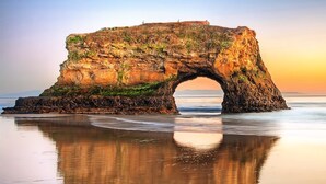 Formação rochosa natural em arco, com vista clara do oceano e um pôr do sol ao fundo.
