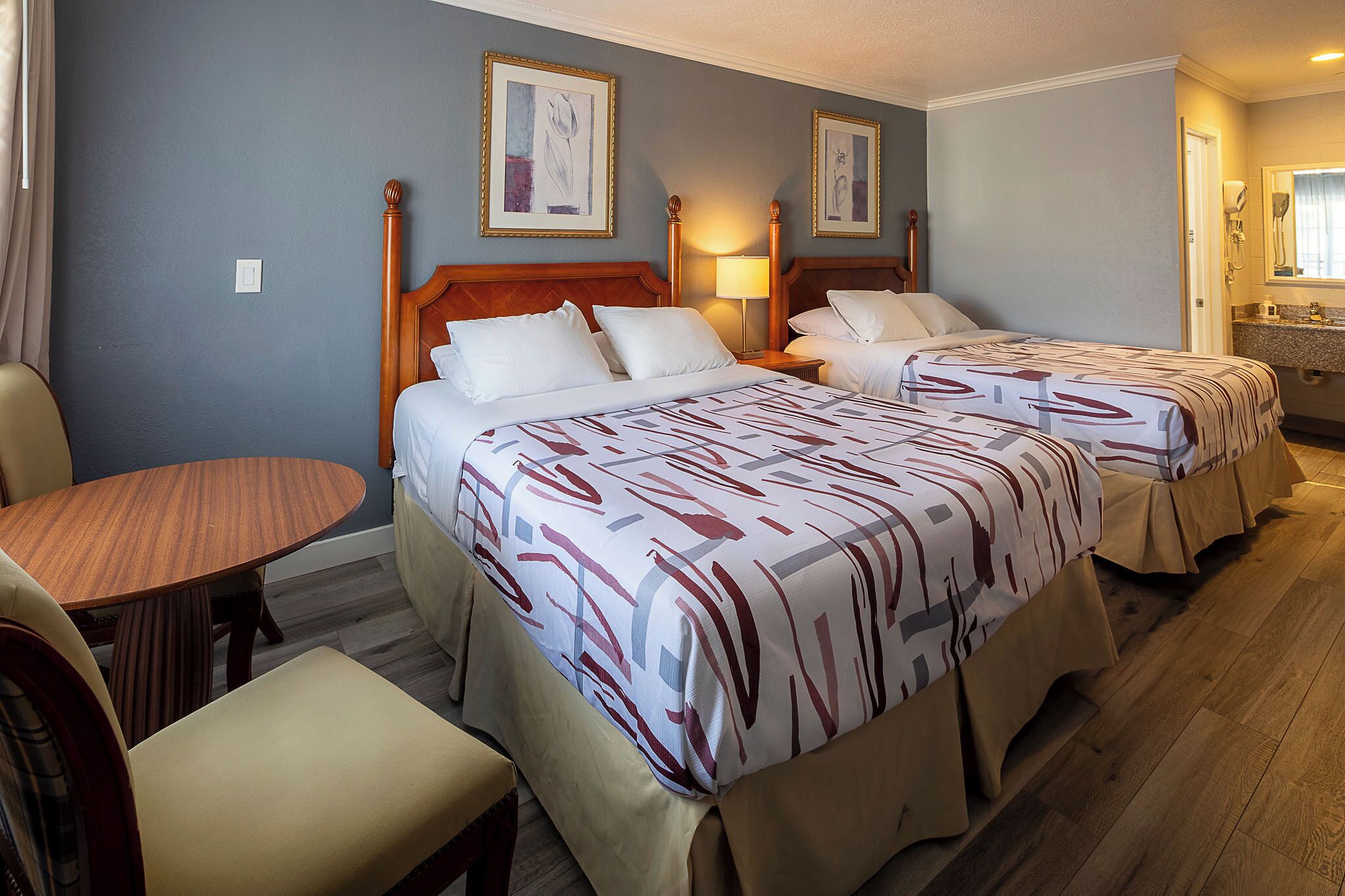 Quarto de hotel com duas camas, uma cabeceira de madeira, uma mesa redonda de madeira e uma cadeira.